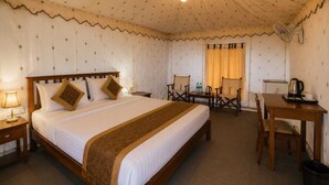 Quarto de hotel com uma cama grande, duas cadeiras, uma mesa pequena, uma escrivaninha, uma chaleira e um ventilador.
