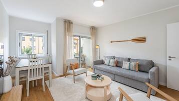 Uma sala de estar moderna com um sofá cinzento, uma mesa de jantar com cadeiras, uma mesa de centro redonda e vista para o exterior pela janela.