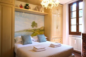 Quarto com cama, travesseiros, pintura de paisagem, lustre e janela.