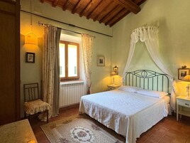 Um quarto com cama, janela com cortinas, mesa de cabeceira com abajur e uma cadeira.