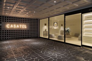 Entrada moderna de hotel com piso de pedra, fachada de vidro e o nome 'CASATEL' iluminado na parede.
