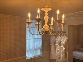 Quarto com lustre, janela com cortinas e cama com colcha branca.