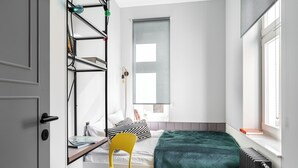 A compact bedroom with a bed, a desk, a chair, a radiator, and a window with blinds.