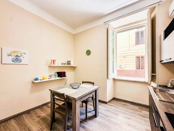 Uma pequena sala de jantar com uma mesa e cadeiras, uma janela com vista para edifícios e uma parede com prateleiras contendo diversos objetos.