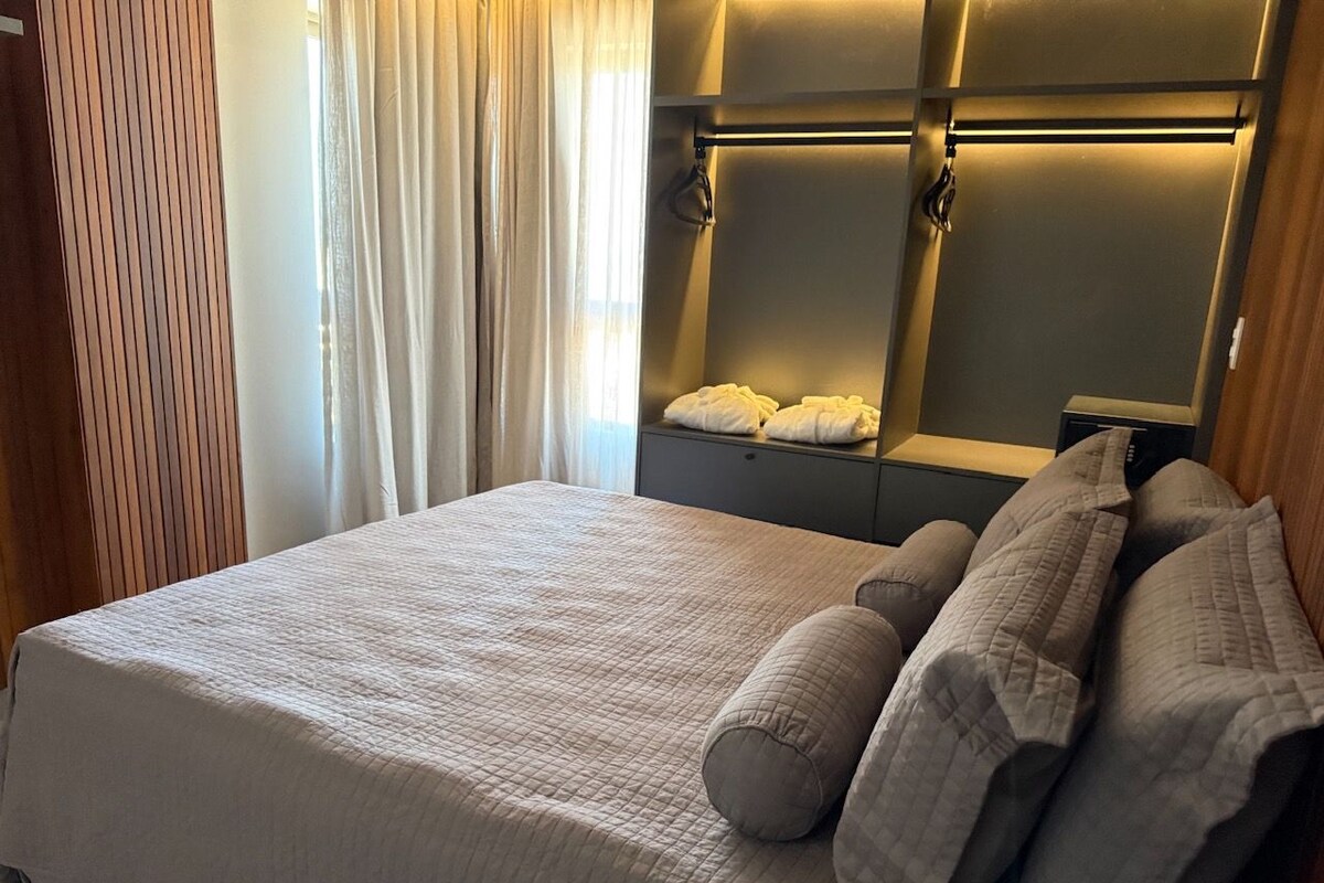 Quarto de hotel com cama, mesa de cabeceira, espelho e janela com cortinas.