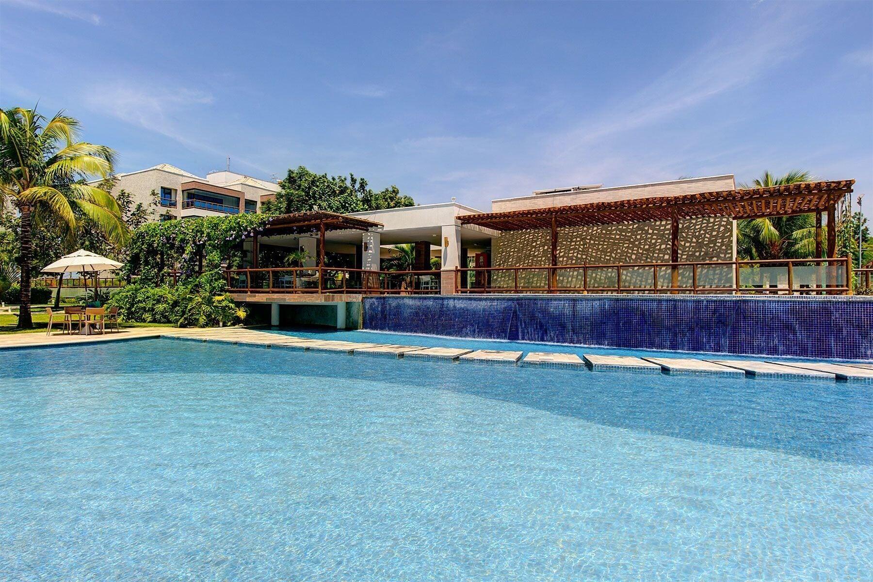 Piscina com superfície de água azul cristalina, rodeada por um deck de madeira e uma área coberta com pérgola.