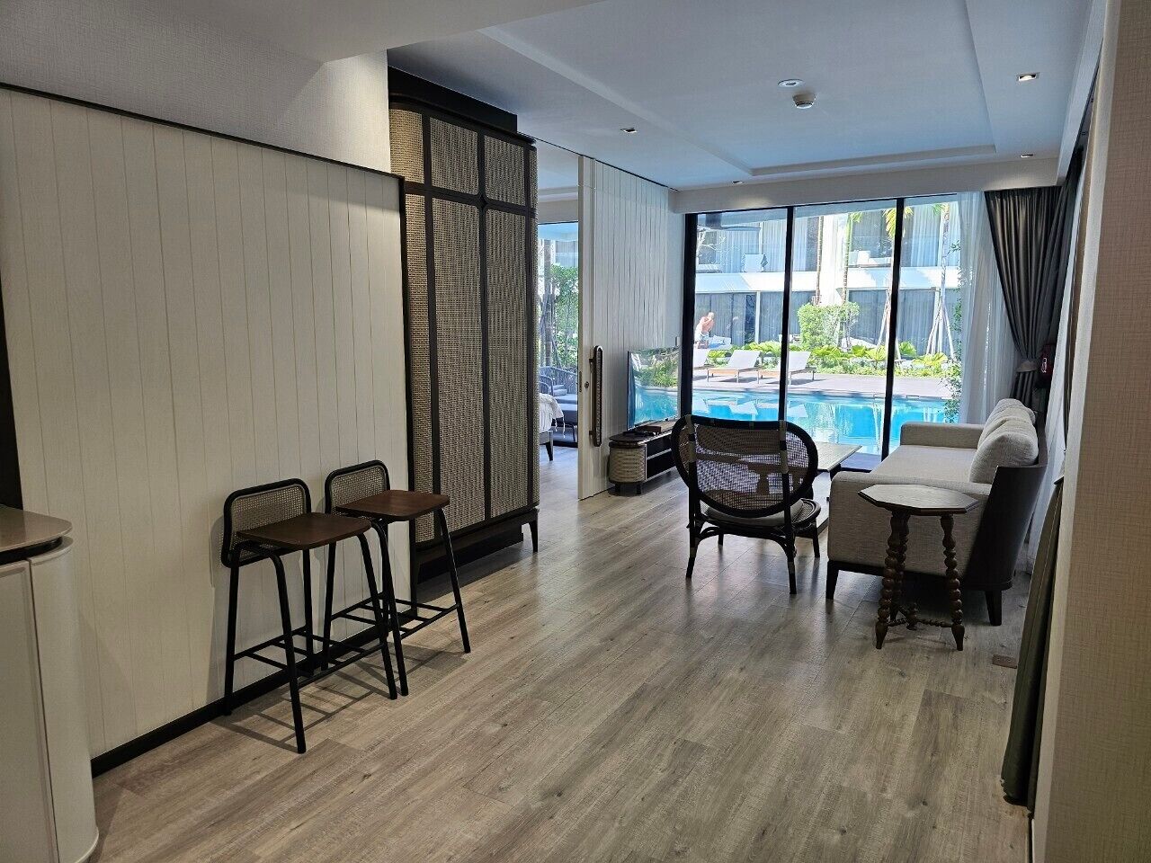 Quarto de hotel moderno com piso de madeira, um sofá, uma mesa de jantar com cadeiras e uma janela grande com vista para uma piscina.
