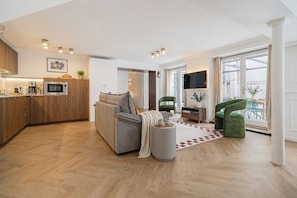 Uma sala de estar moderna com um sofá cinzento, uma poltrona verde, uma cozinha com armários de madeira e uma janela ampla.