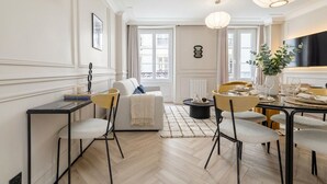 Uma sala de estar moderna com sofá, mesa de jantar, cadeiras e mesa de centro. Há uma janela com vista para edifícios.