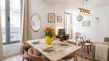 Uma sala de jantar com uma mesa posta para quatro pessoas, um vaso amarelo com flores e uma televisão ao fundo.