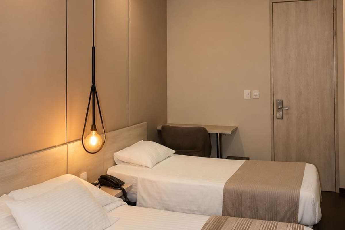 Quarto de hotel com duas camas, uma mesa, uma cadeira e uma porta.