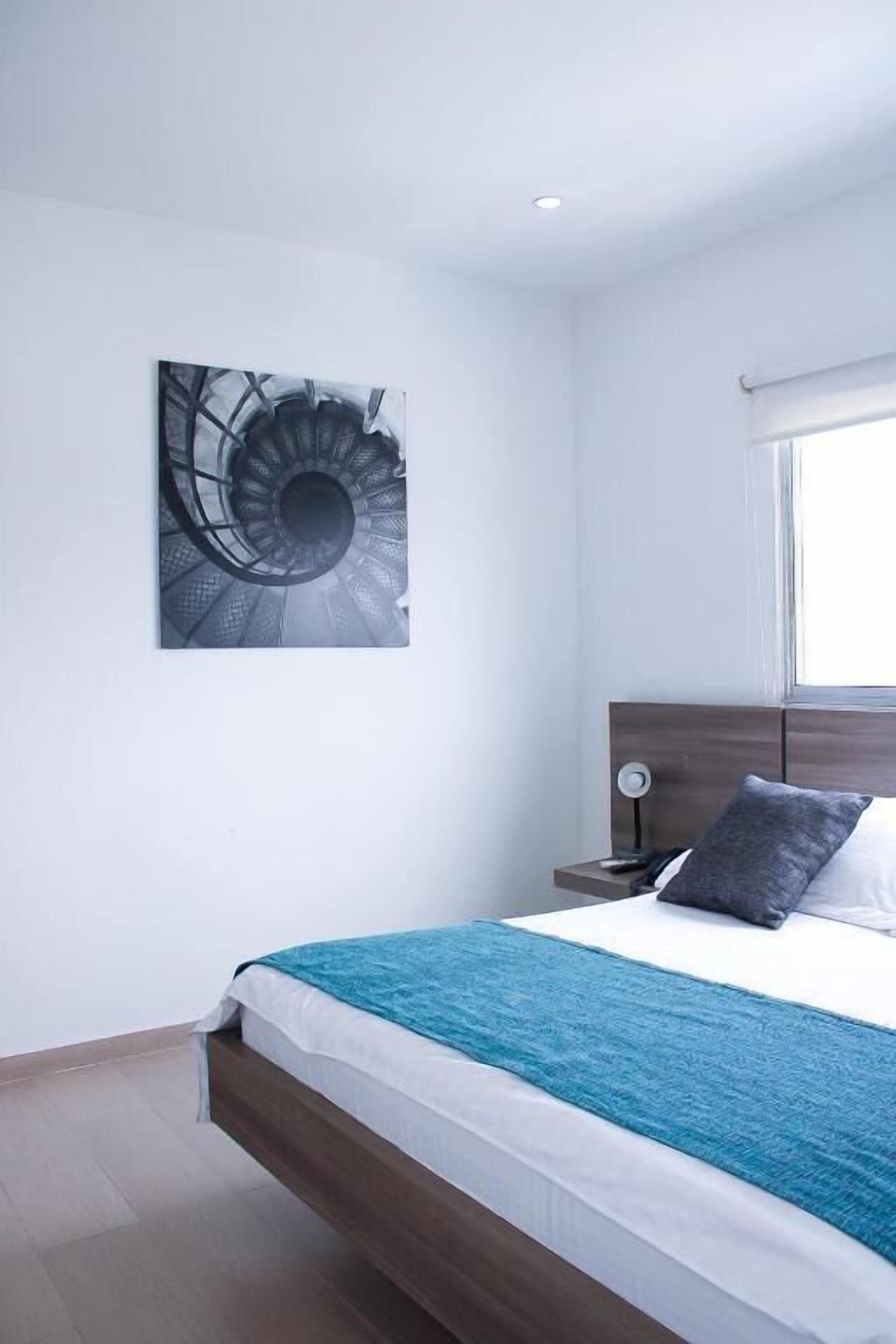 Um quarto com cama, mesa de cabeceira, uma obra de arte na parede e uma janela.