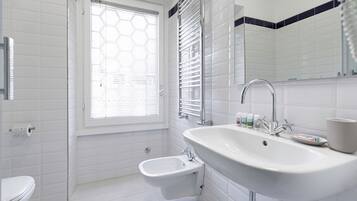Casa de banho moderna com lavatório branco, bidé e sanita. Tem uma janela com persianas, espelho e porta-toalhas.