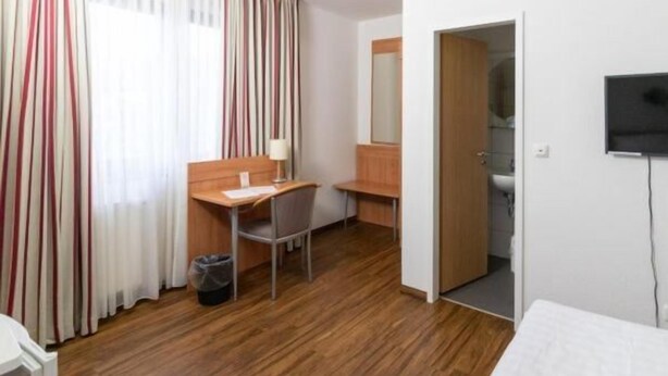 Quarto de hotel com piso de madeira, uma escrivaninha com cadeira, uma cama, televisão e banheiro visível por uma porta aberta.