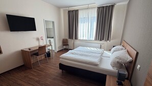 Quarto de hotel com cama, escrivaninha, cadeira, televisão fixada na parede e janela com cortinas.