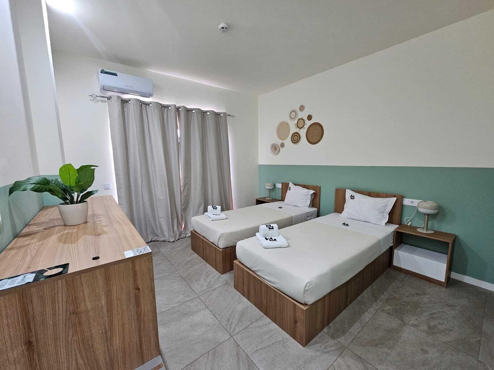 Quarto de hotel com duas camas, uma escrivaninha, um vaso de planta e ar condicionado fixado na parede.