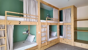 Quarto beliche com três níveis, cada um com um número. Cortinas nas janelas, um ventilador na parede e uma estante com abajur.