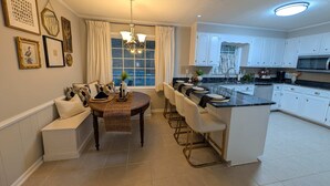 Uma cozinha moderna com armários brancos, uma área de refeições com mesa e cadeiras redondas, e uma janela grande com cortinas.