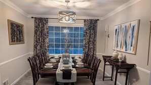 Uma sala de jantar com uma mesa comprida posta para seis pessoas, com cadeiras, um arranjo de flores no centro e uma janela com cortinas.