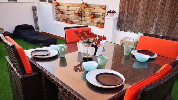 Uma área de refeições com mesa de vidro, cadeiras de vime com almofadas laranja e um painel de madeira decorado com plantas em vasos.
