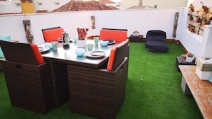Um terraço no telhado com uma mesa para quatro pessoas, rodeado por cadeiras de vime com almofadas laranja.