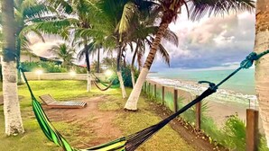 Uma rede amarrada entre palmeiras, com vista para uma praia, um cercado e um deck de madeira.