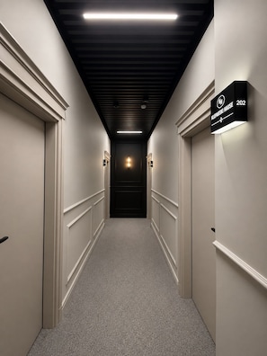 Um corredor com teto preto, paredes brancas e piso coberto por carpete. Há duas portas de cada lado e uma placa sobre a porta à direita.