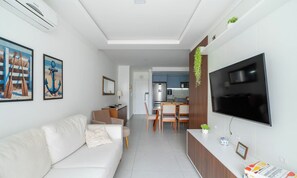 Uma sala de estar moderna com um sofá branco, uma TV de tela plana na parede e uma área de jantar com cadeiras de madeira e uma pequena mesa.
