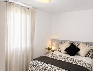 Quarto com cama, cortinas, mesa de cabeceira com abajur e luminária na parede.