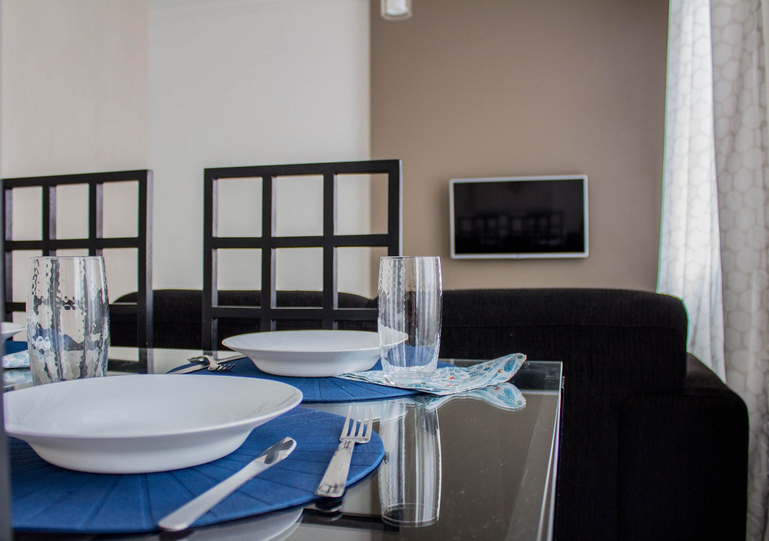 Mesa posta com pratos, talheres e copos, com uma cadeira preta e um quadro na parede.