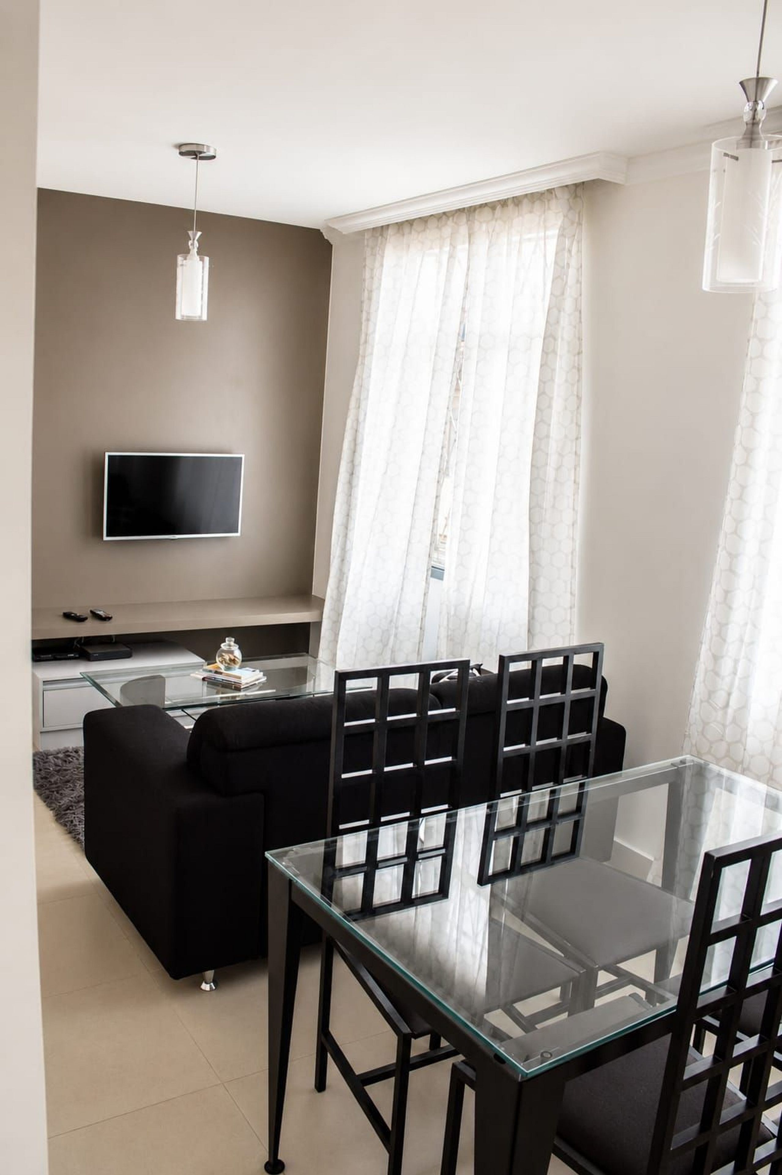 Uma sala de estar moderna com mesa de jantar de vidro, cadeiras pretas, sofá preto, televisão de tela plana e uma mesa de centro com livros e um vaso.