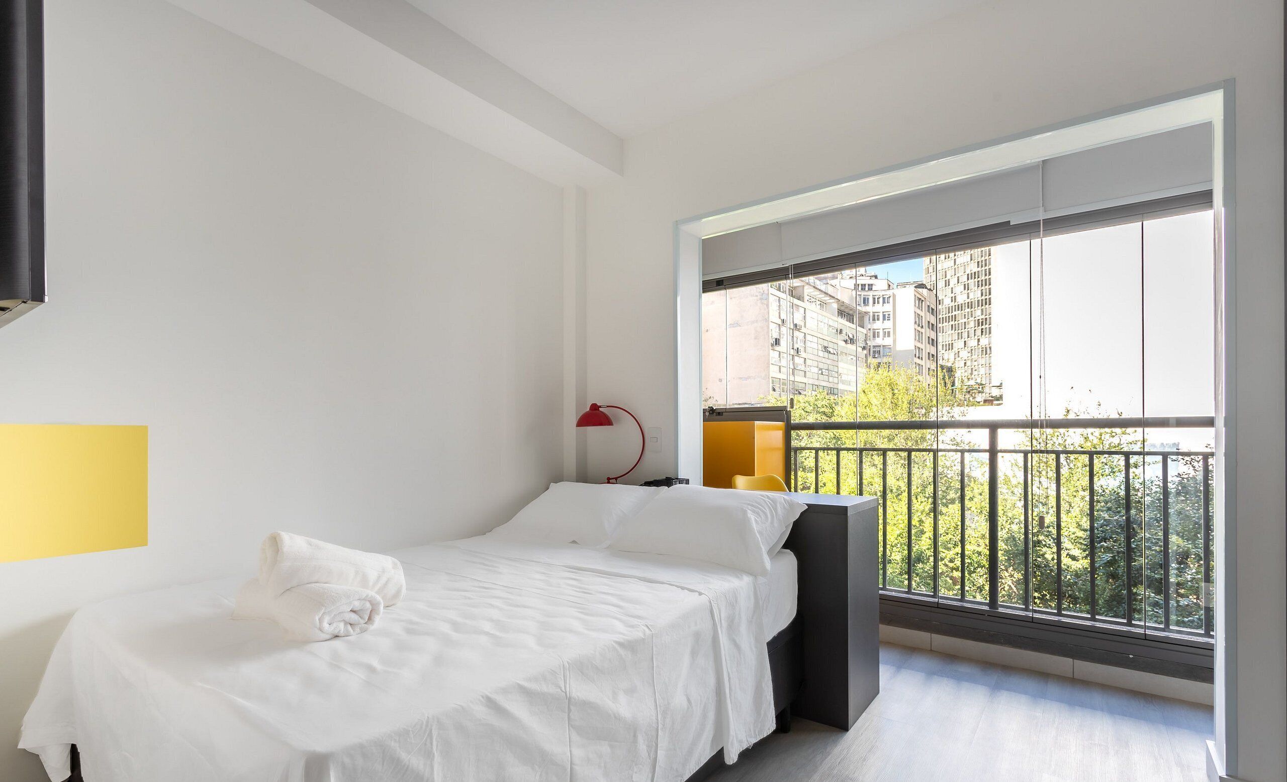 Quarto de hotel minimalista com uma cama, uma luminária de mesa vermelha e uma grande janela com vista para a cidade.