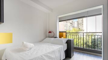 Quarto de hotel minimalista com uma cama, uma luminária de mesa vermelha e uma grande janela com vista para a cidade.