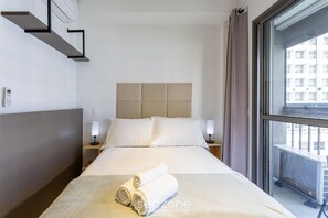 Quarto de hotel com cama, duas mesas de cabeceira, encosto e janela com vista para prédios.