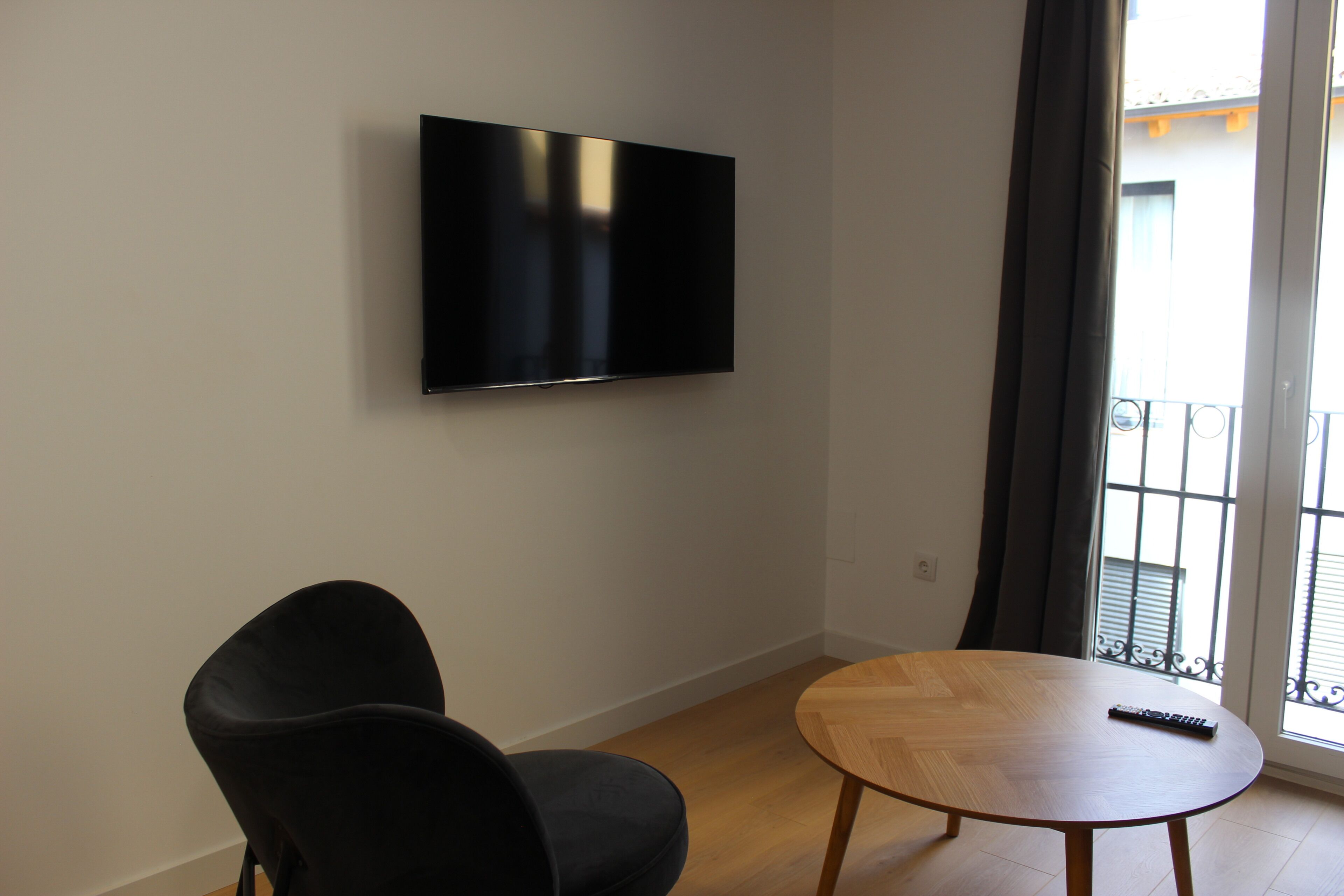Quarto com televisão de tela plana na parede, uma mesa de madeira redonda e uma cadeira preta.