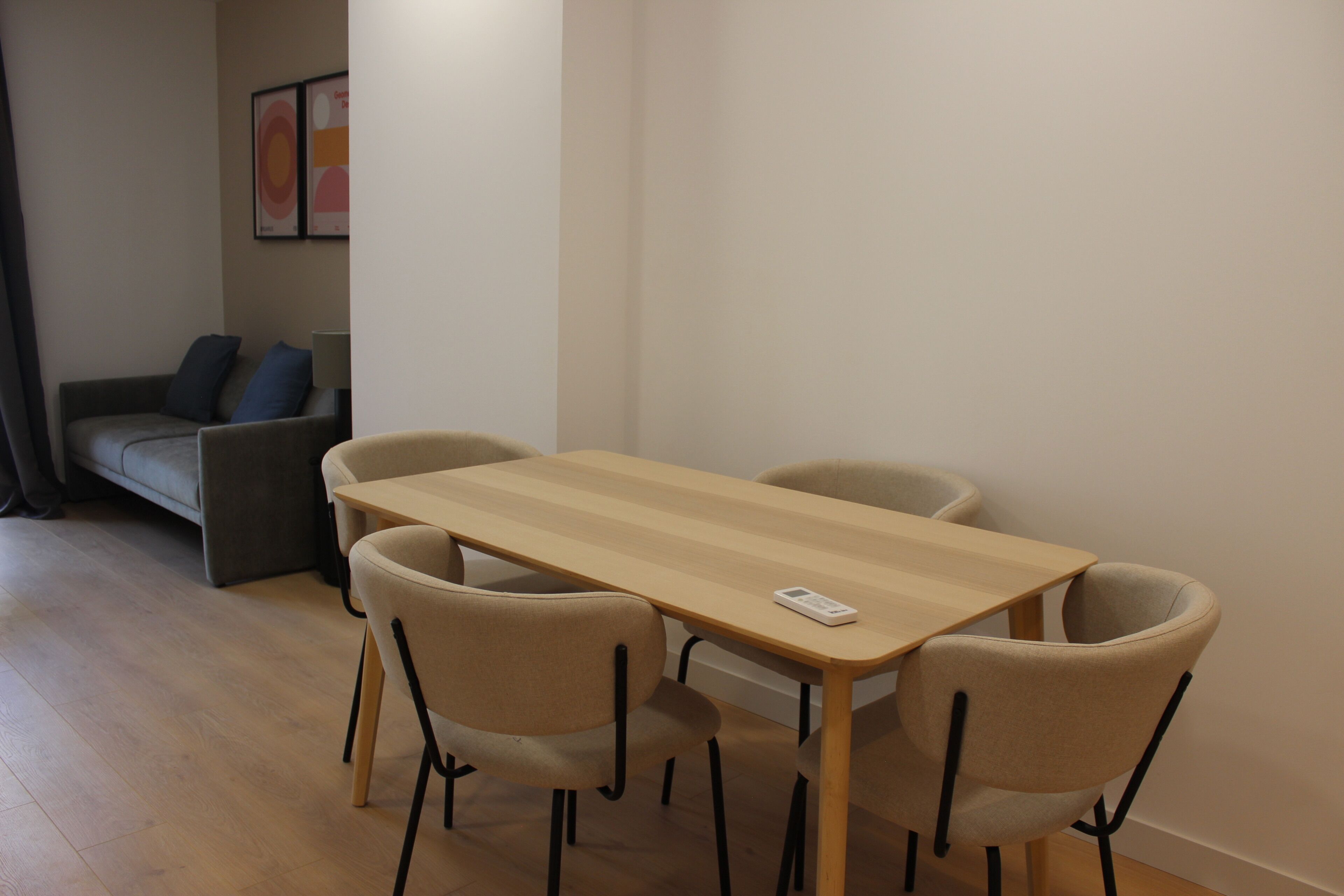 Um quarto com uma mesa de madeira e quatro cadeiras, um sofá cinzento e uma luminária.