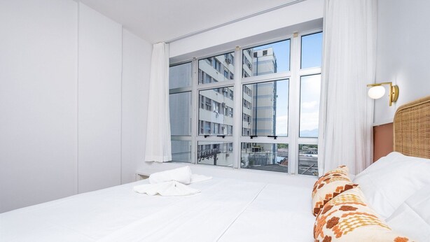 Quarto de hotel com uma janela grande, cama com roupa de cama branca e vista para um edifício.