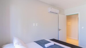 Quarto de hotel com uma cama de solteiro, mesa de cabeceira, ar condicionado fixado na parede e uma porta que dá para outro quarto.