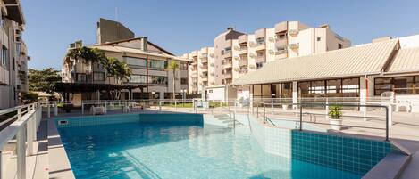 Uma piscina com um deck revestido, rodeada por edifícios altos com varandas e uma área coberta com assentos.