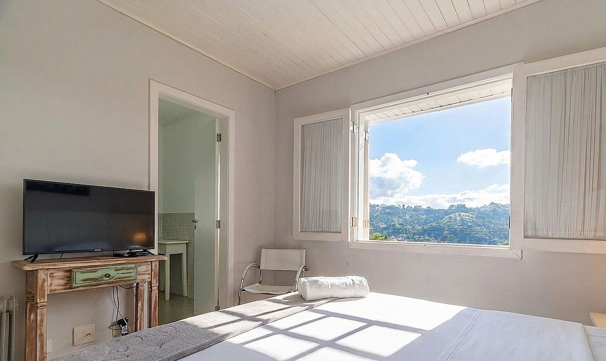 Quarto de hotel com uma cama, televisão sobre um suporte de madeira, banheiro com chuveiro e janela com vista para as montanhas.