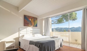 Quarto com cama, mesa de cabeceira, quadro e vista para o exterior.