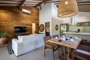 Uma sala de estar moderna com um sofá branco, uma mesa de jantar posta para quatro pessoas e uma cozinha com geladeira e luminária pendente.