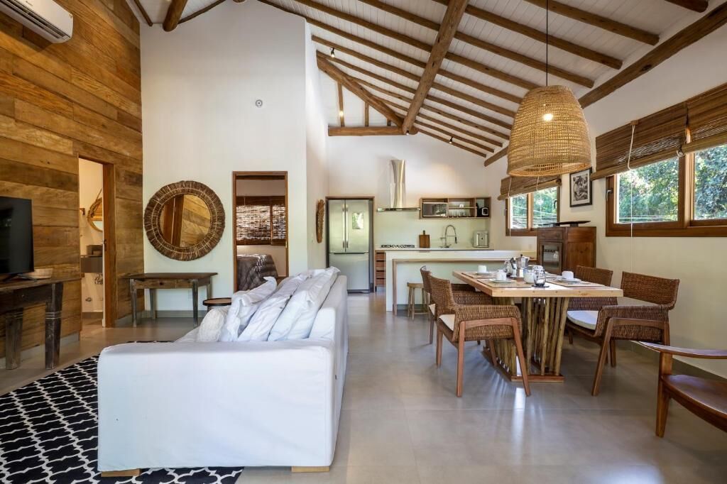 Uma sala de estar ampla com sofá, mesa de jantar e cadeiras, cozinha ao fundo e uma grande janela que traz luz natural.