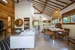 Uma sala de estar ampla com sofá, mesa de jantar e cadeiras, cozinha ao fundo e uma grande janela que traz luz natural.