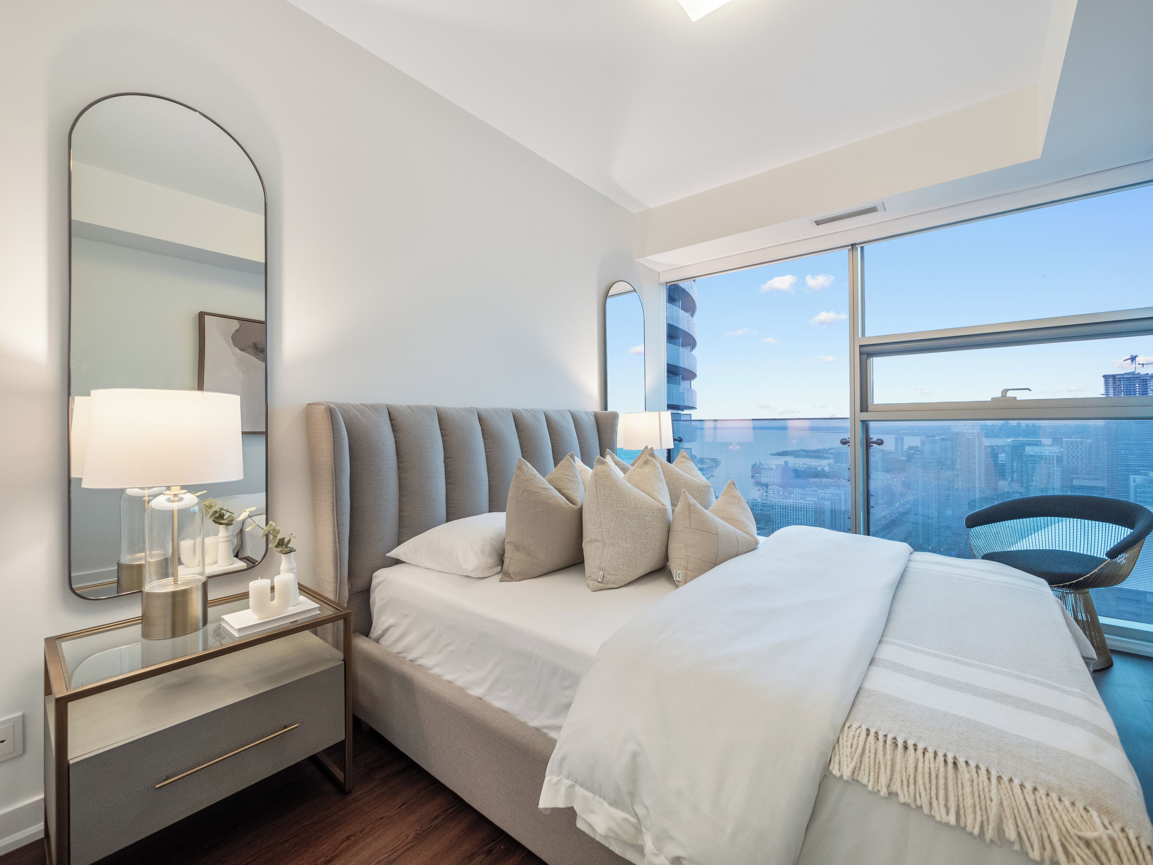 Quarto moderno com uma cama grande, mesa de cabeceira, espelho e vista da cidade através de amplas janelas.