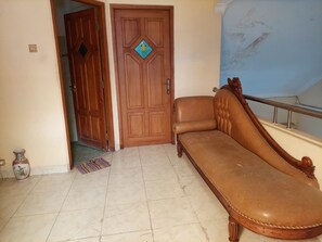 Um quarto com um sofá marrom, uma porta de madeira com um janelão em formato de diamante e chão de azulejos.