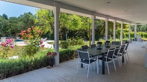 Um pátio externo com mesa e cadeiras, cercado por vegetação e vista para uma piscina.