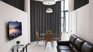 Uma sala de estar moderna com um sofá de couro, uma mesa de jantar com cadeiras, uma televisão de tela plana na parede e uma janela grande com cortinas.
