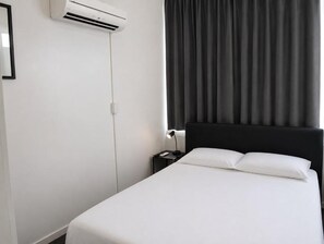 Quarto de hotel com cama, mesa de cabeceira, luminária, ar condicionado e janela com cortinas.