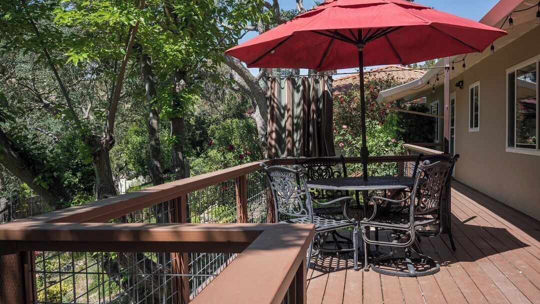 Um deck de madeira com uma mesa e cadeiras, um guarda-sol vermelho, e vista para árvores e uma casa.